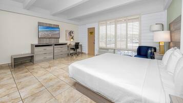 Quarto de hotel moderno com uma cama grande, uma mesa com cadeira, TV e vista da cidade por grandes janelas.
