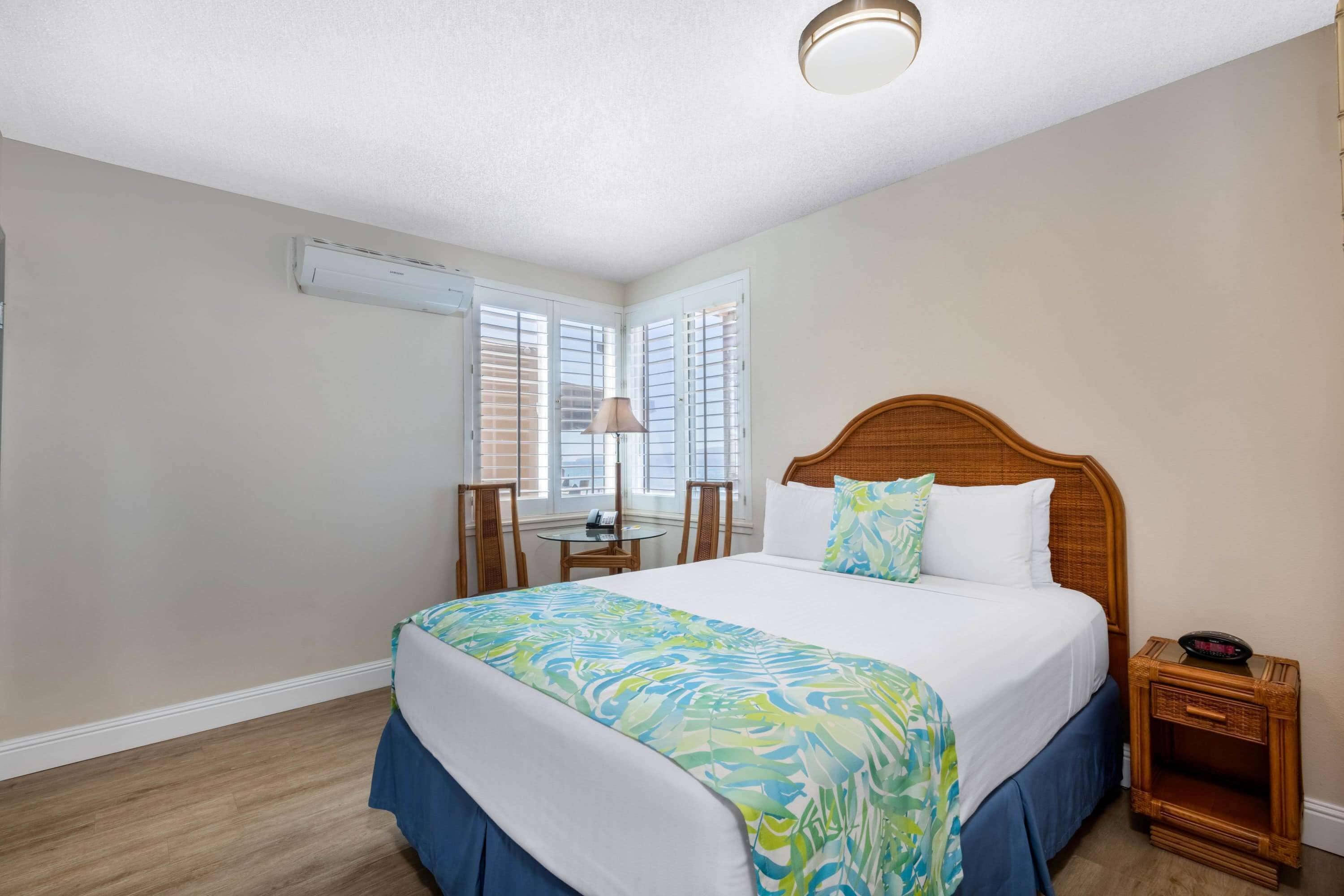 Quarto de hotel com uma cama grande, cabeceira de madeira, mesa de cabeceira e janela com persianas.