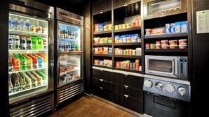 Uma estação de alimentos e bebidas com serviço livre, incluindo dispensadores de bebidas refrigeradas, prateleiras com lanches e um forno micro-ondas.
