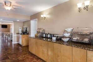 Espaço de café da manhã de hotel, com balcão exibindo diversos alimentos, ventilador de teto e iluminação embutida nas paredes.
