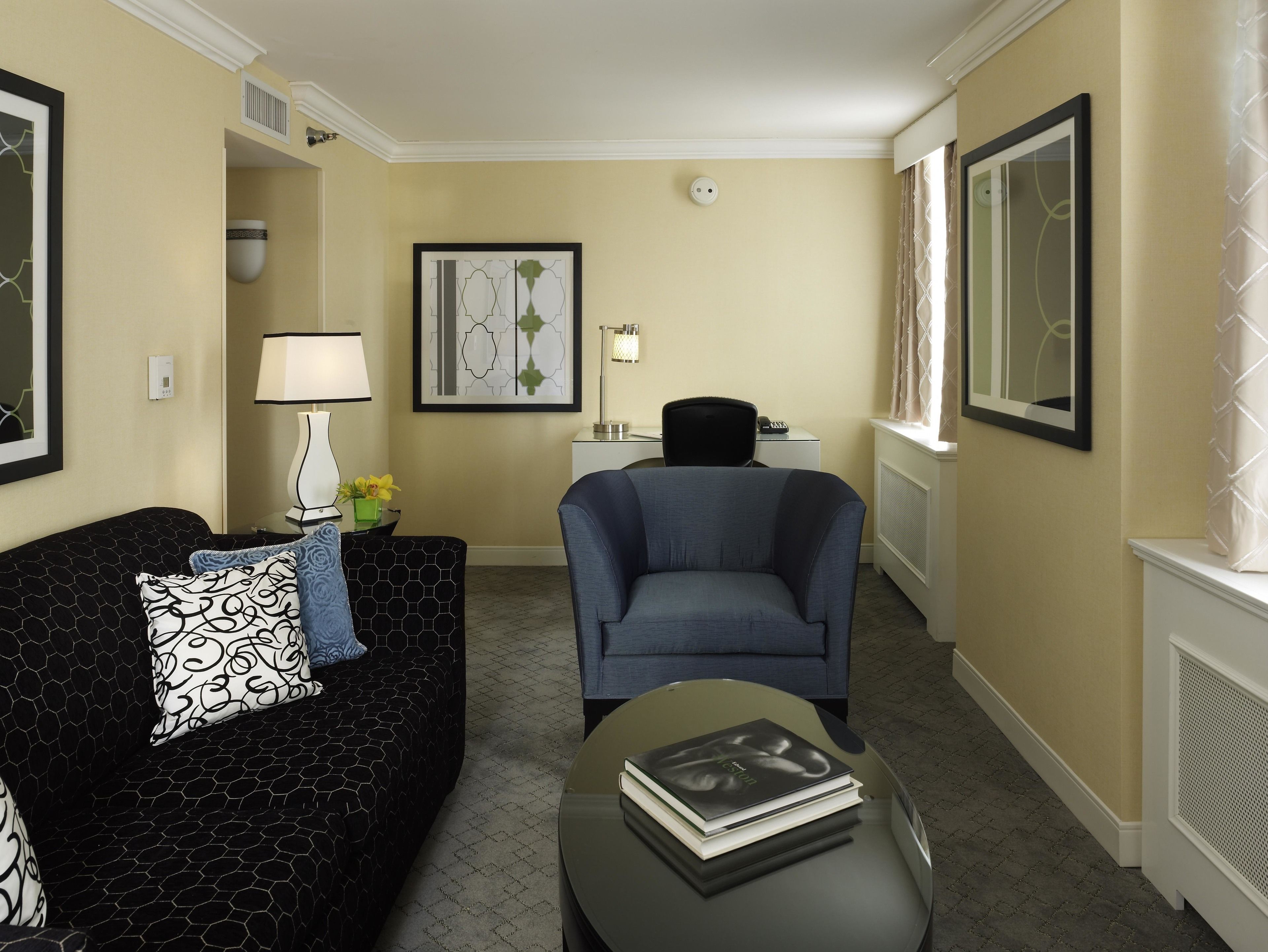 Sala de estar com um sofá estampado preto, uma poltrona azul e uma mesa de centro com livros. Há um quadro na parede, um abajur e uma janela com cortinas.