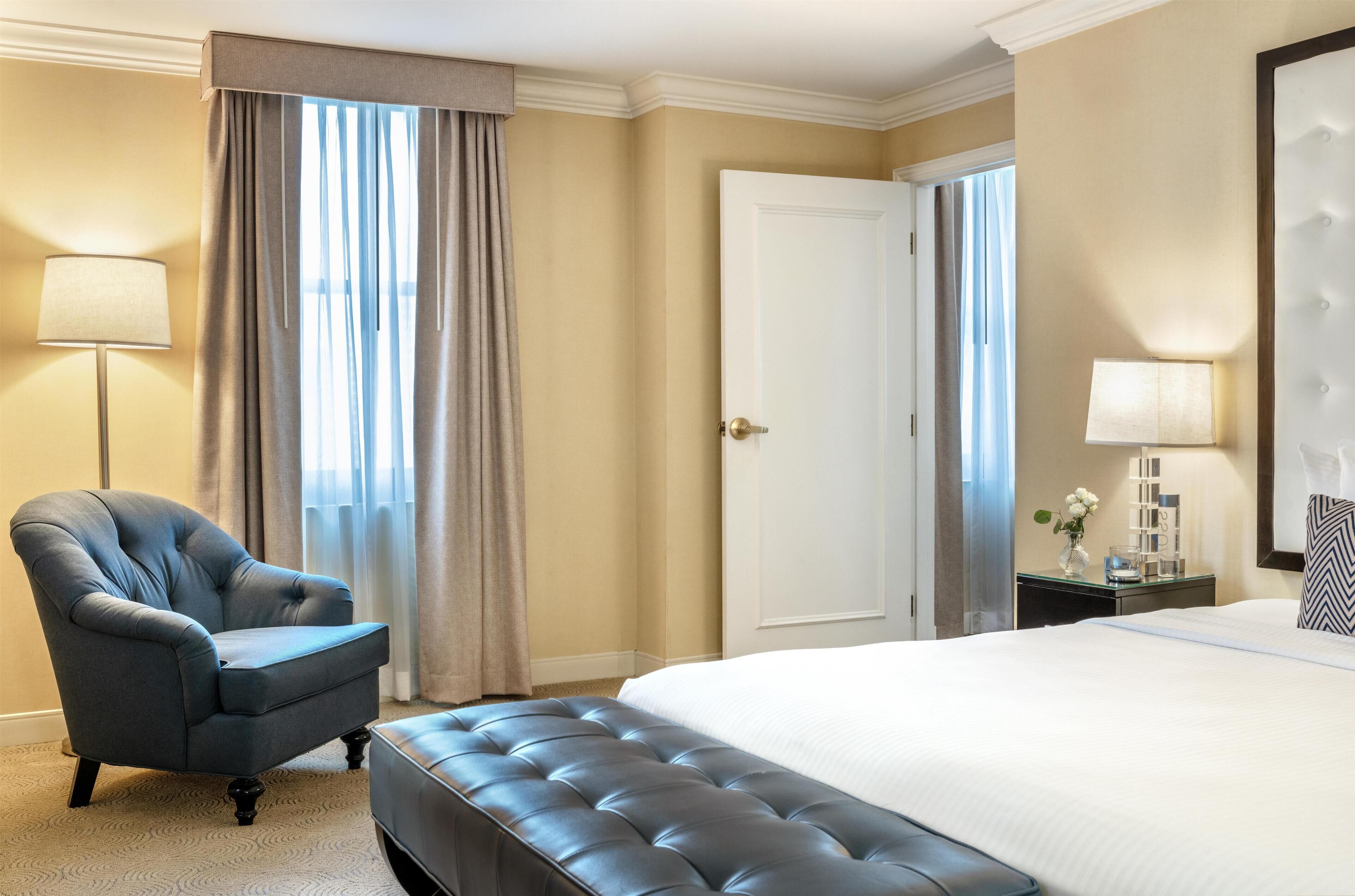 Quarto de hotel com uma cama grande, uma cadeira, uma mesa de cabeceira com abajur e uma porta.