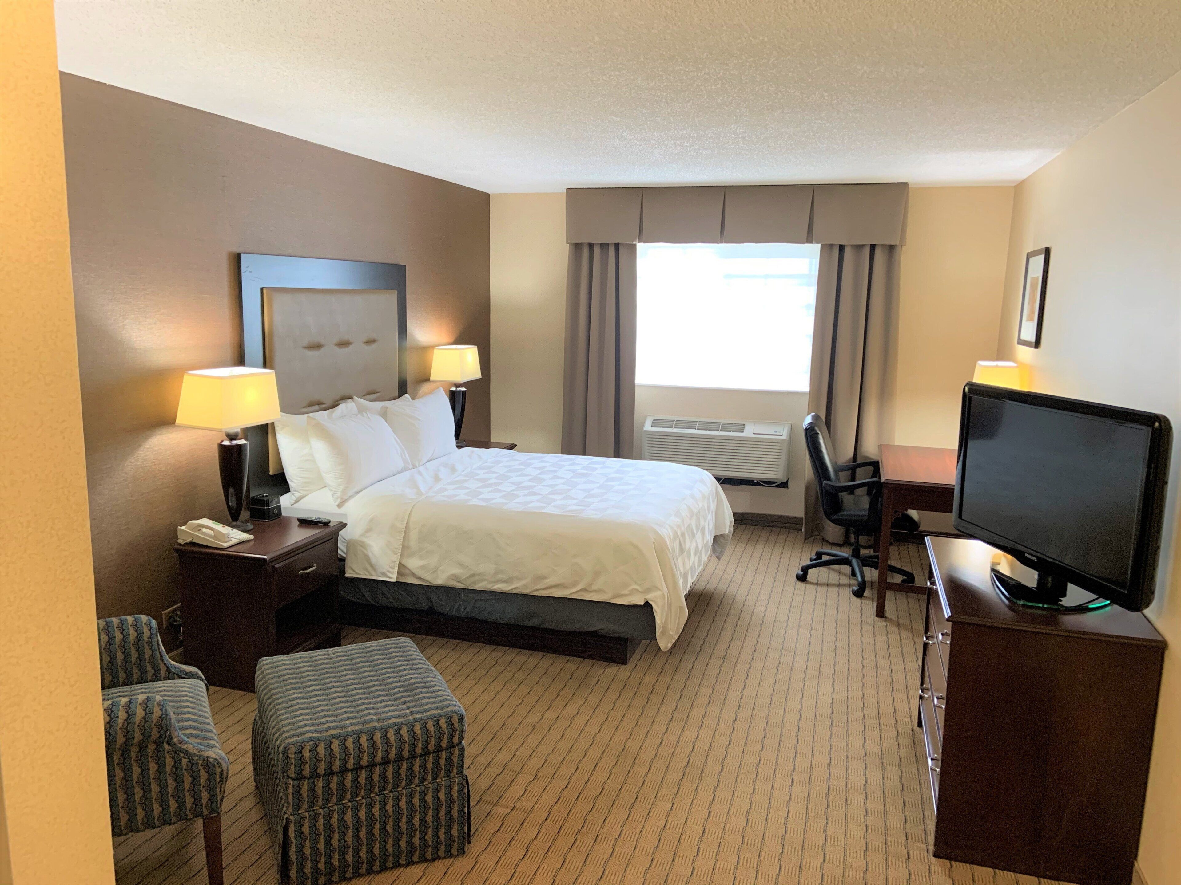 Quarto de hotel com cama, duas luminárias de cabeceira, escrivaninha com computador, cadeira, banquinho e televisão.