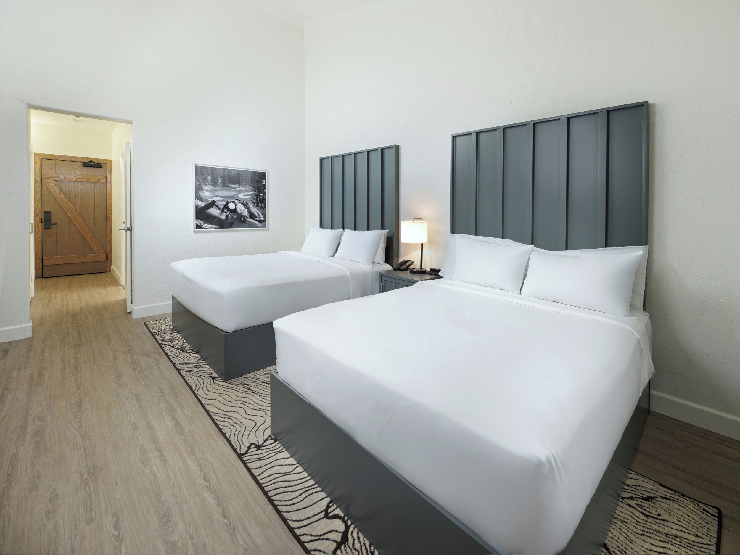 Quarto de hotel com duas camas, uma porta de madeira, um quadro na parede e um abajur.