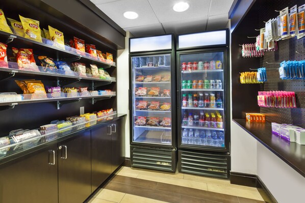 Uma área de lanches e bebidas, com prateleiras de salgadinhos, um refrigerador com bebidas e uma parede com itens pendurados.