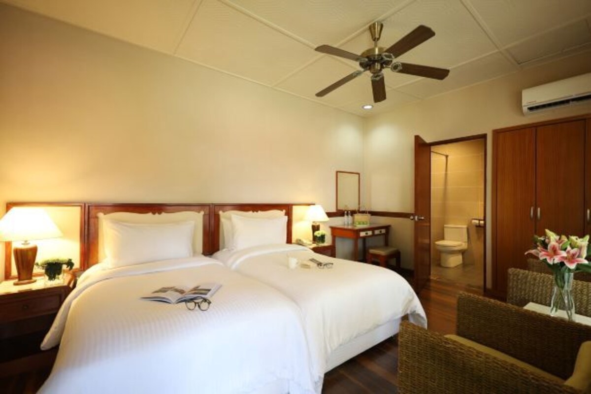 Quarto de hotel com uma cama grande, uma escrivaninha com cadeira, ventoinha no teto, e banheiro com vaso sanitário e chuveiro.