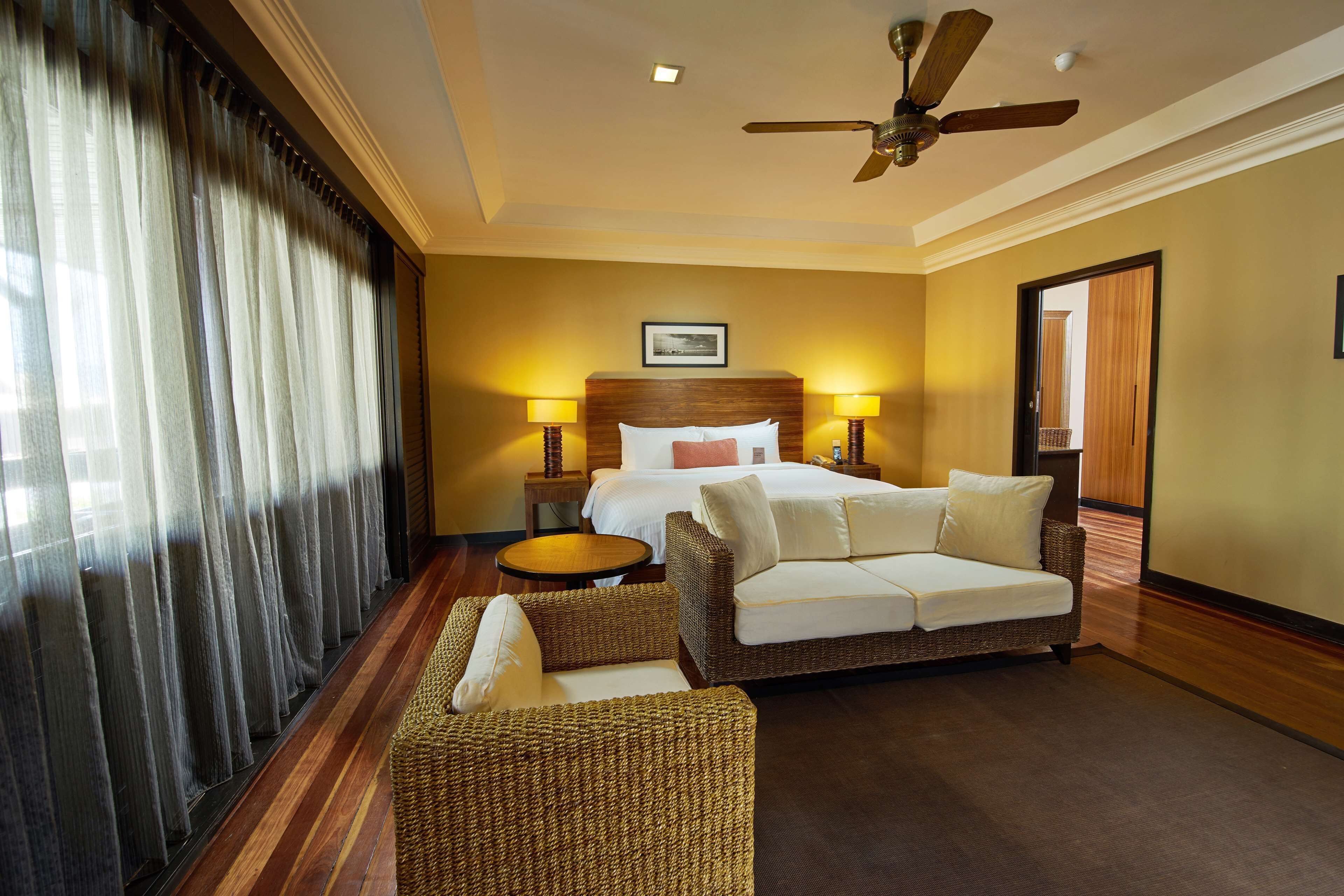 Quarto de hotel com uma cama, duas mesas de cabeceira com luminárias, um sofá, uma cadeira, um ventilador de teto e uma janela grande com cortinas.
