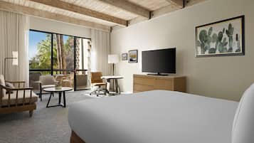 Quarto de hotel com uma cama grande, televisão de tela plana, escrivaninha e uma cadeira. Há uma janela com cortinas e vista para palmeiras.