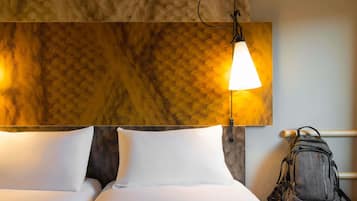 Quarto de hotel com cama, almofadas, abajur, parede com textura e uma mochila num criado-mudo.