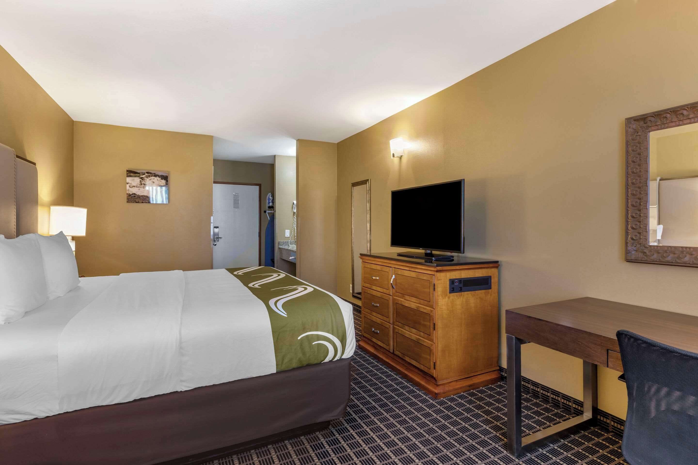 Quarto de hotel com cama, penteadeira de madeira, escrivaninha, televisão e espelho.
