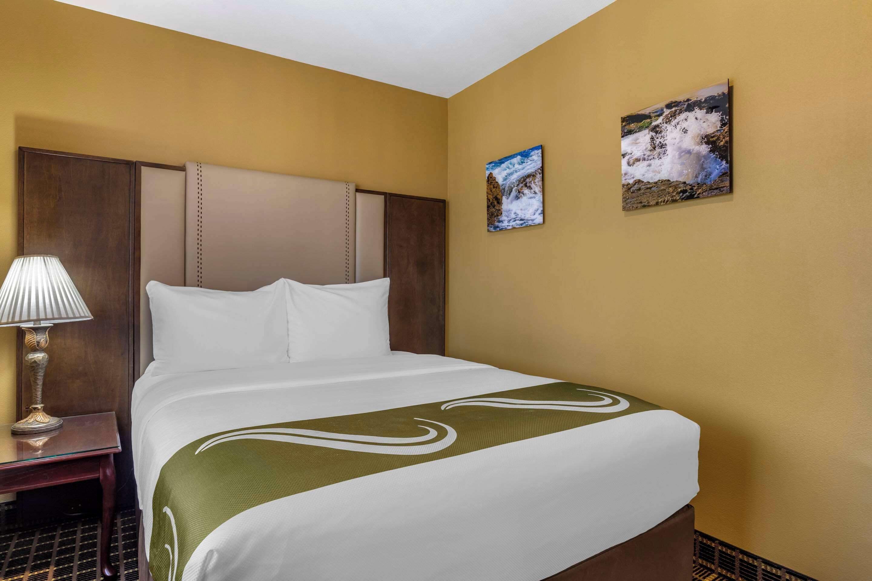 Quarto de hotel com uma cama, uma mesinha de cabeceira com luminária e duas imagens emolduradas na parede.
