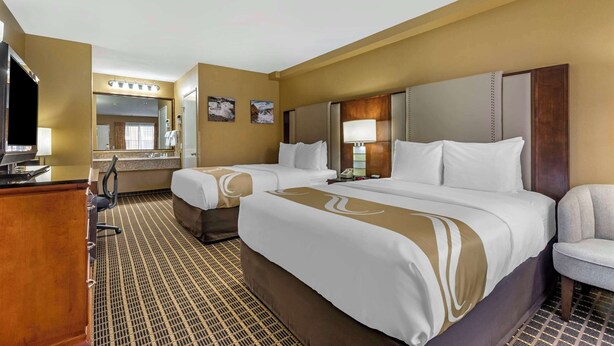Quarto de hotel com duas camas, uma escrivaninha, uma cadeira, uma televisão e um banheiro visível por uma porta aberta.