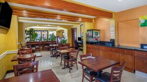 Restaurante com mesas e cadeiras de madeira, área de cozinha e plantas em vasos.
