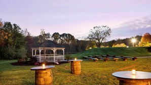Um gazebo em uma área gramada com barris de madeira e bancos, cercado por árvores e uma cerca branca.