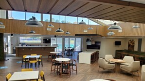 Amplo espaço interno com diversas opções de assentos, incluindo mesas e cadeiras, uma ilha central com banquetas e uma área de cozinha com balcão e eletrodomésticos.