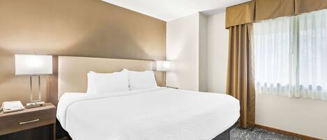 Quarto de hotel com uma cama grande, uma mesinha de cabeceira com abajur e uma janela com cortinas.