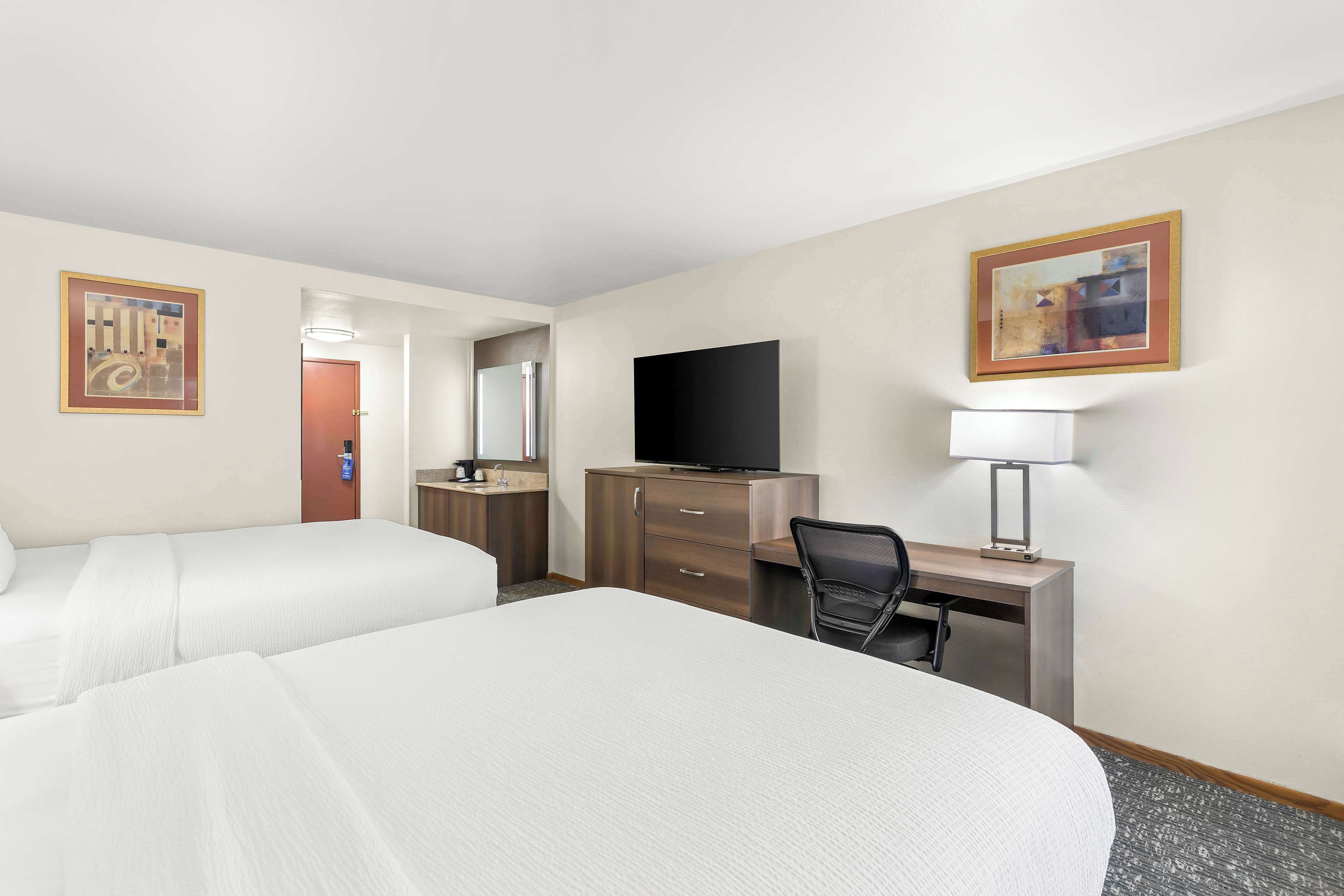 Quarto de hotel com duas camas, uma escrivaninha com cadeira, televisão e dois quadros nas paredes.