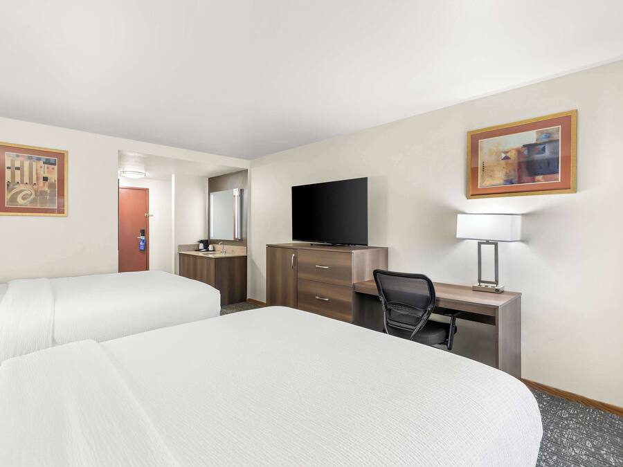 Quarto de hotel com duas camas, uma escrivaninha com cadeira, televisão e dois quadros nas paredes.