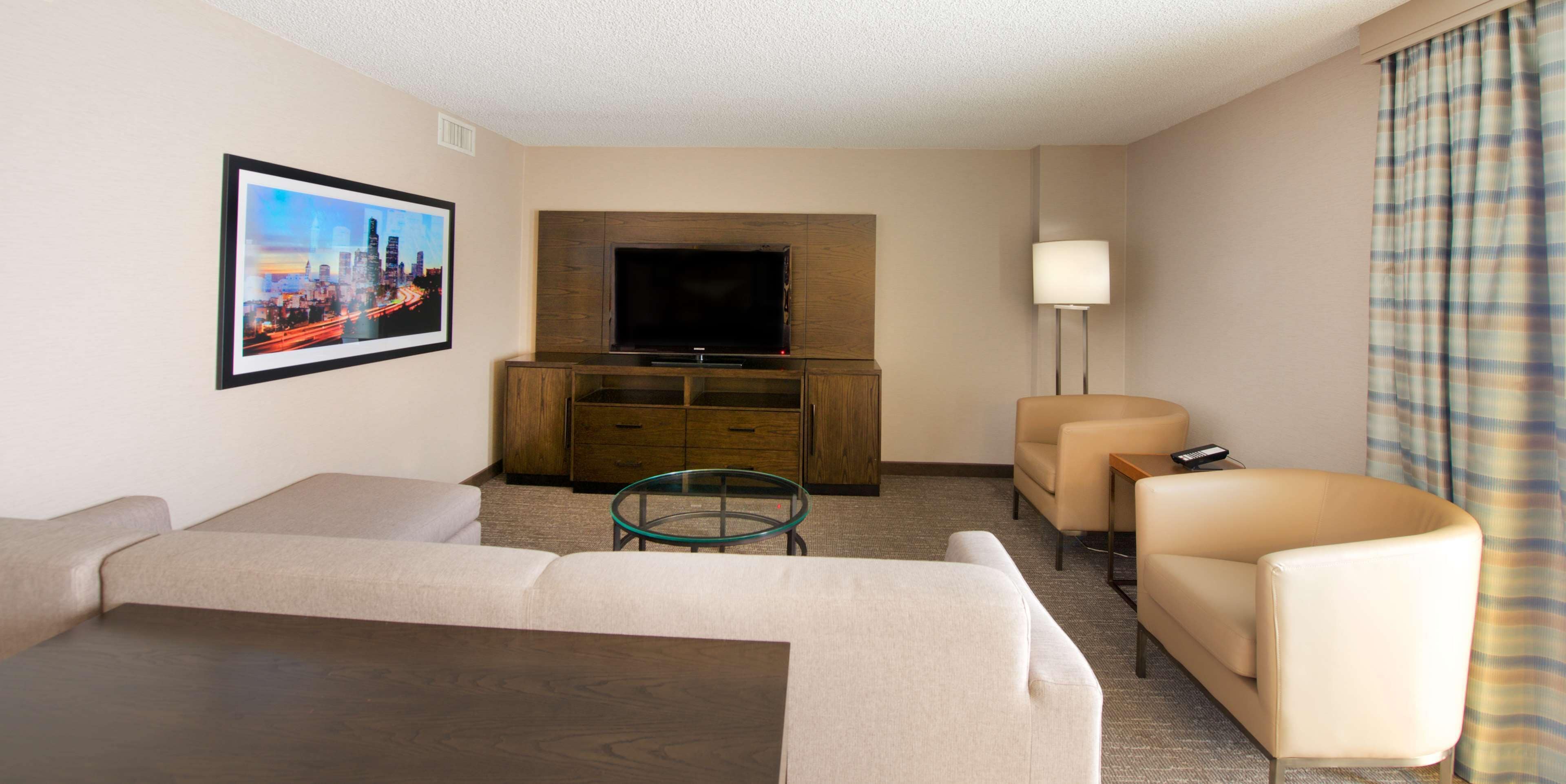 Quarto de hotel com uma televisão de tela grande, um sofá, uma cadeira, uma mesinha e um abajur.