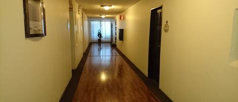 Corredor de hotel com piso de madeira, um quadro emoldurado à esquerda e uma porta com número à direita.