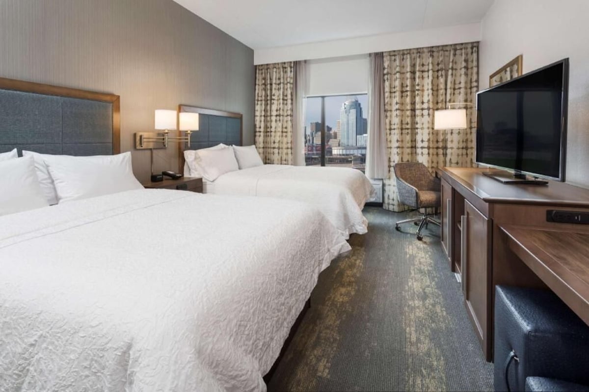 Quarto de hotel com duas camas, uma escrivaninha com televisão, uma cadeira e vista para a cidade.
