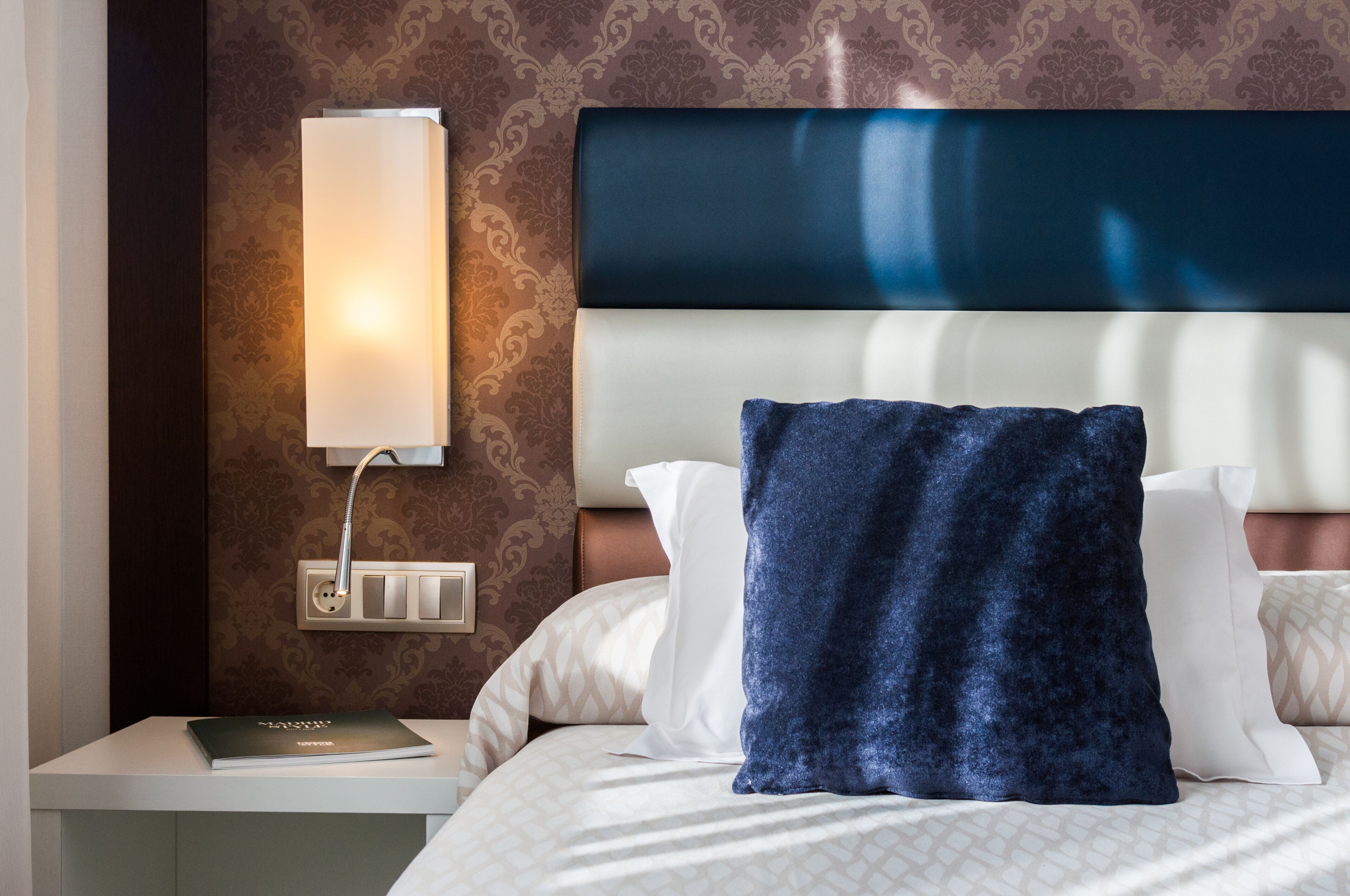 Uma cama bem arrumada com um travesseiro azul, uma luminária de cabeceira e um livro em uma mesa de cabeceira.