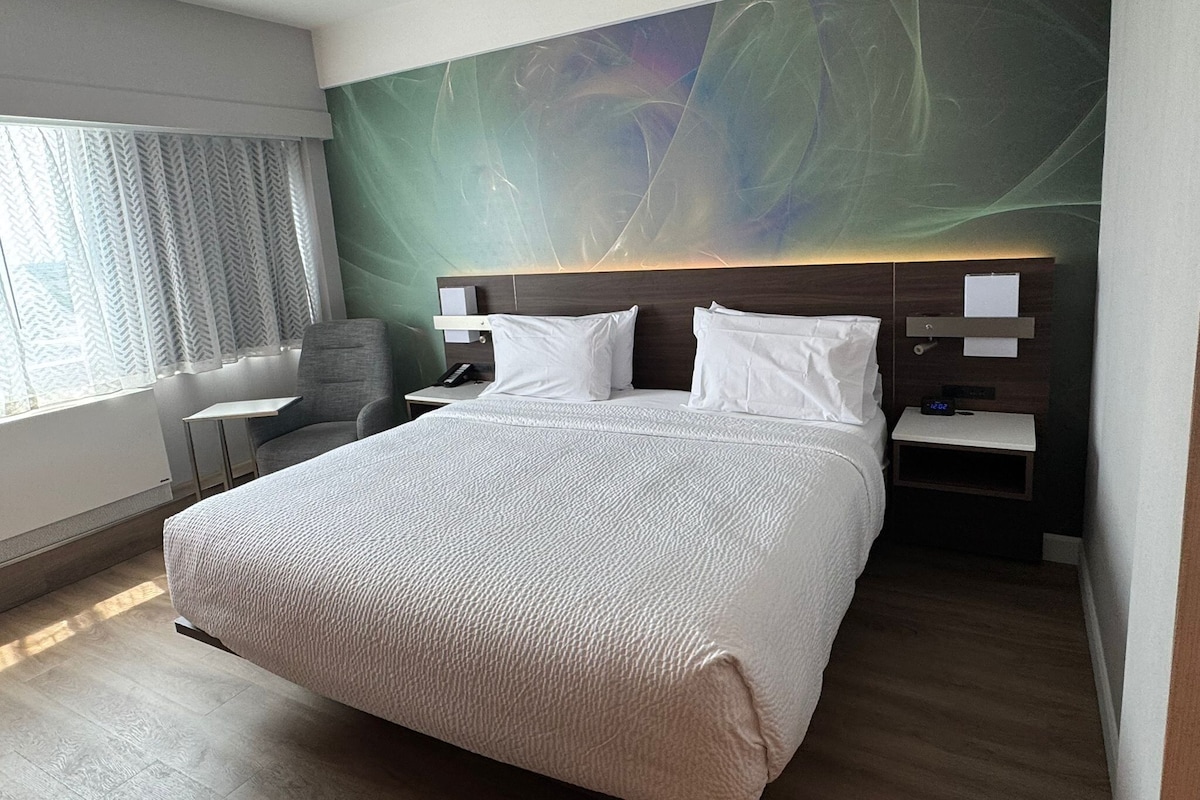 Quarto de hotel moderno com uma cama grande, duas mesas de cabeceira, uma cadeira e vista para o exterior através de cortinas translúcidas.