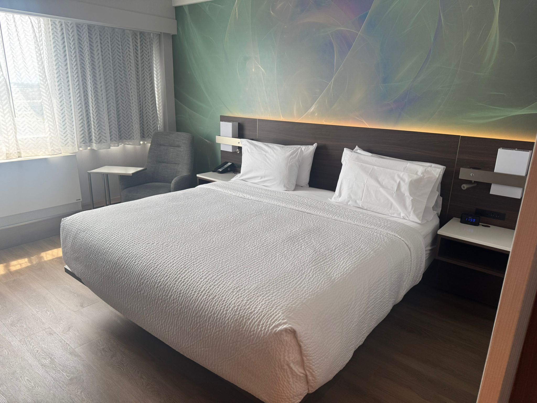 Quarto de hotel moderno com uma cama grande, uma mesa de cabeceira, uma cadeira e uma parede com arte abstrata.
