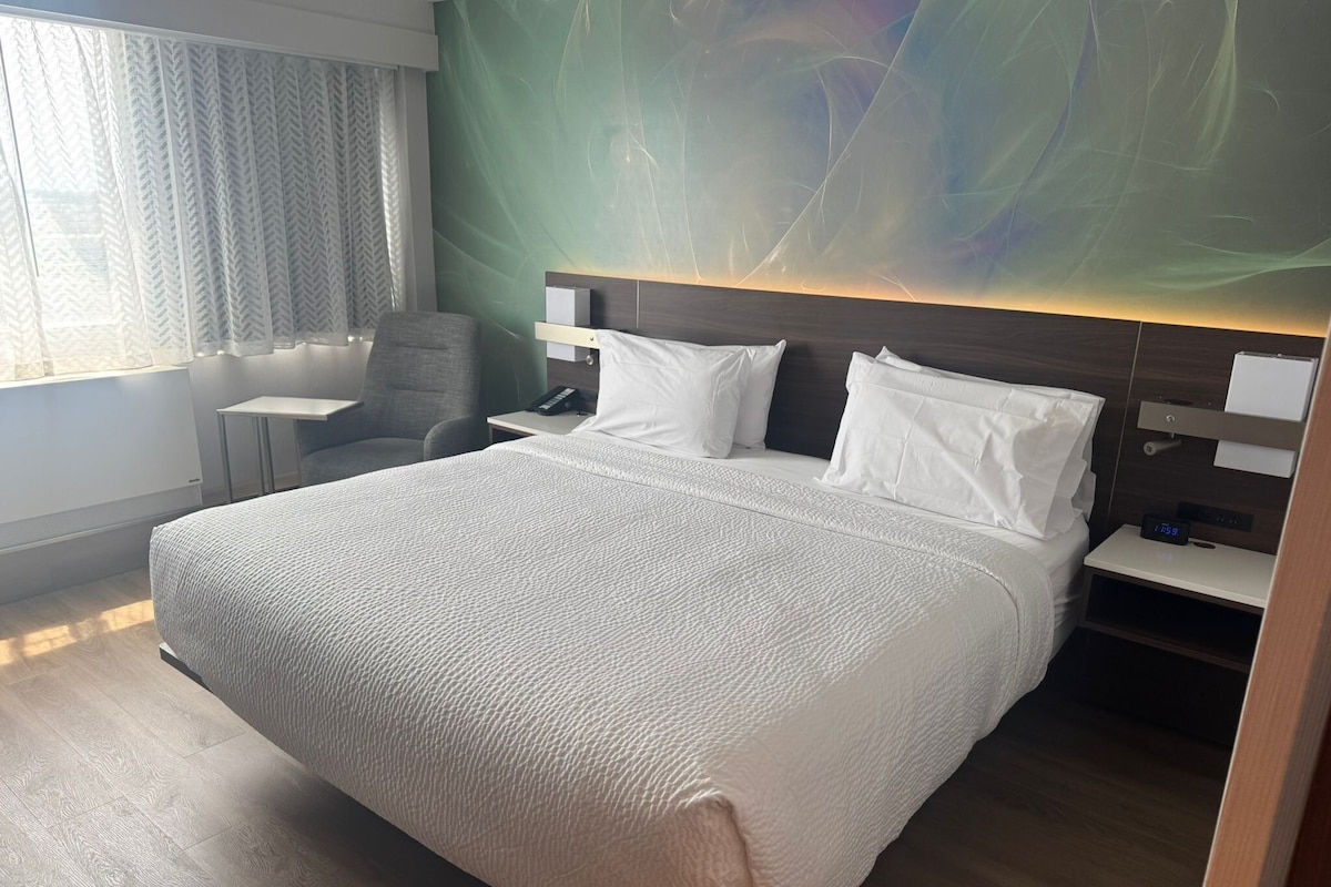 Quarto de hotel moderno com uma cama grande, uma mesinha de cabeceira, uma cadeira e uma parede com arte abstrata.