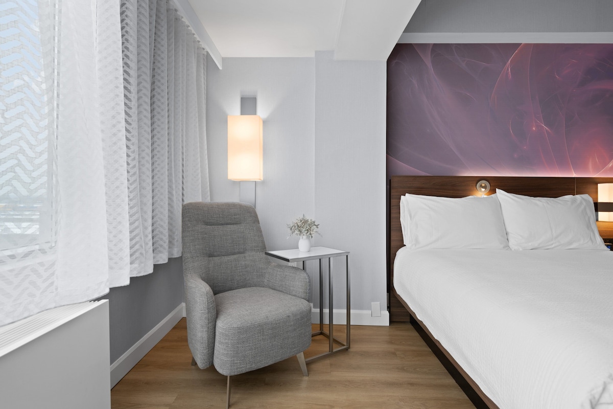 Quarto de hotel com cama, uma poltrona cinzenta, uma mesa pequena com abajur e um objeto decorativo na parede.
