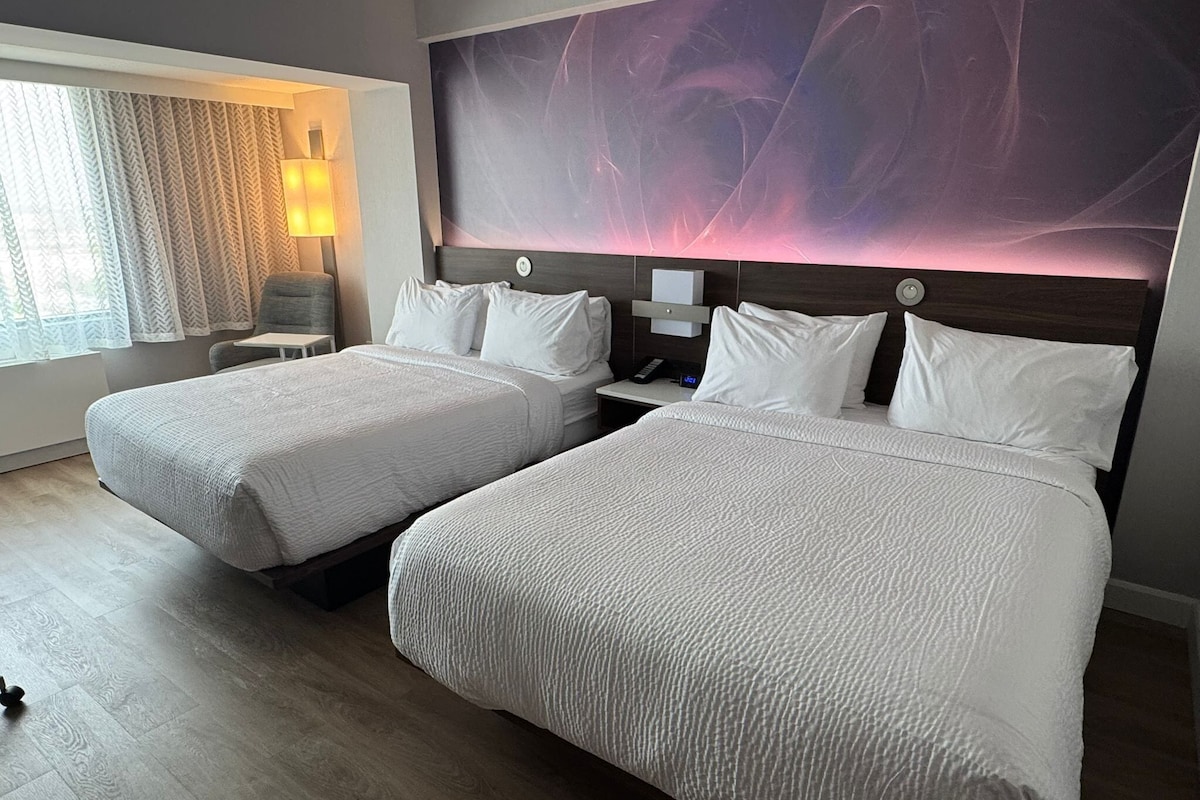 Quarto de hotel com duas camas, uma mesa de cabeceira, uma luminária e um quadro abstrato grande na parede.
