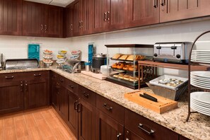 Cozinha com armários de madeira, bancadas de granito, torradeira, display de pães e área de buffet com diversos alimentos.