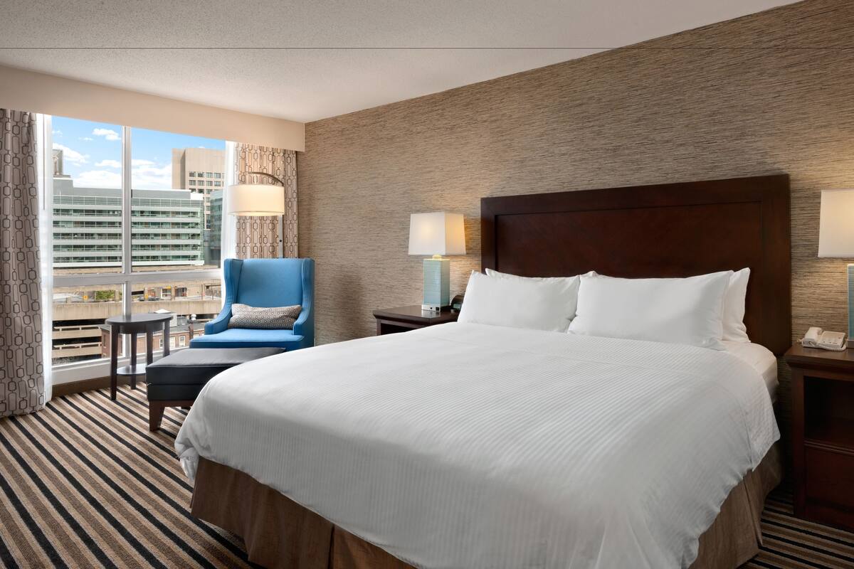 Quarto de hotel com uma cama grande, duas luminárias de cabeceira, uma mesa de apoio, uma cadeira, vista para a cidade e uma janela com cortinas.