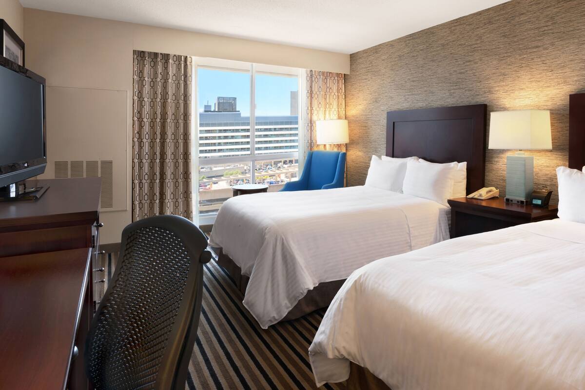 Quarto de hotel com duas camas, uma escrivaninha com televisão, uma cadeira e vista para a cidade.