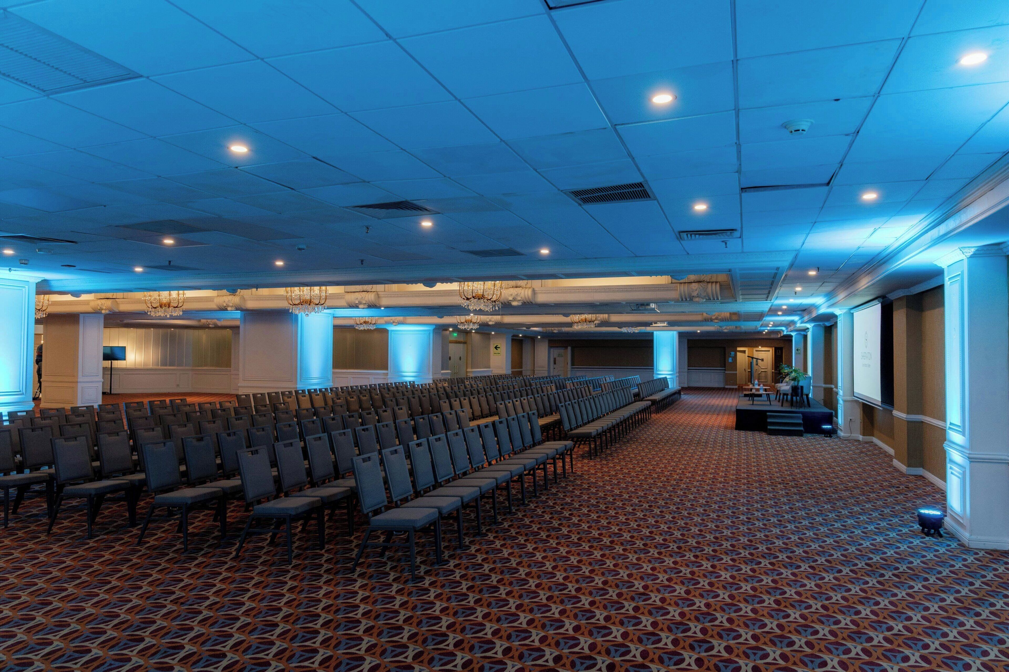 Uma sala de conferências ampla, com fileiras de cadeiras, carpete estampado e teto iluminado em azul.