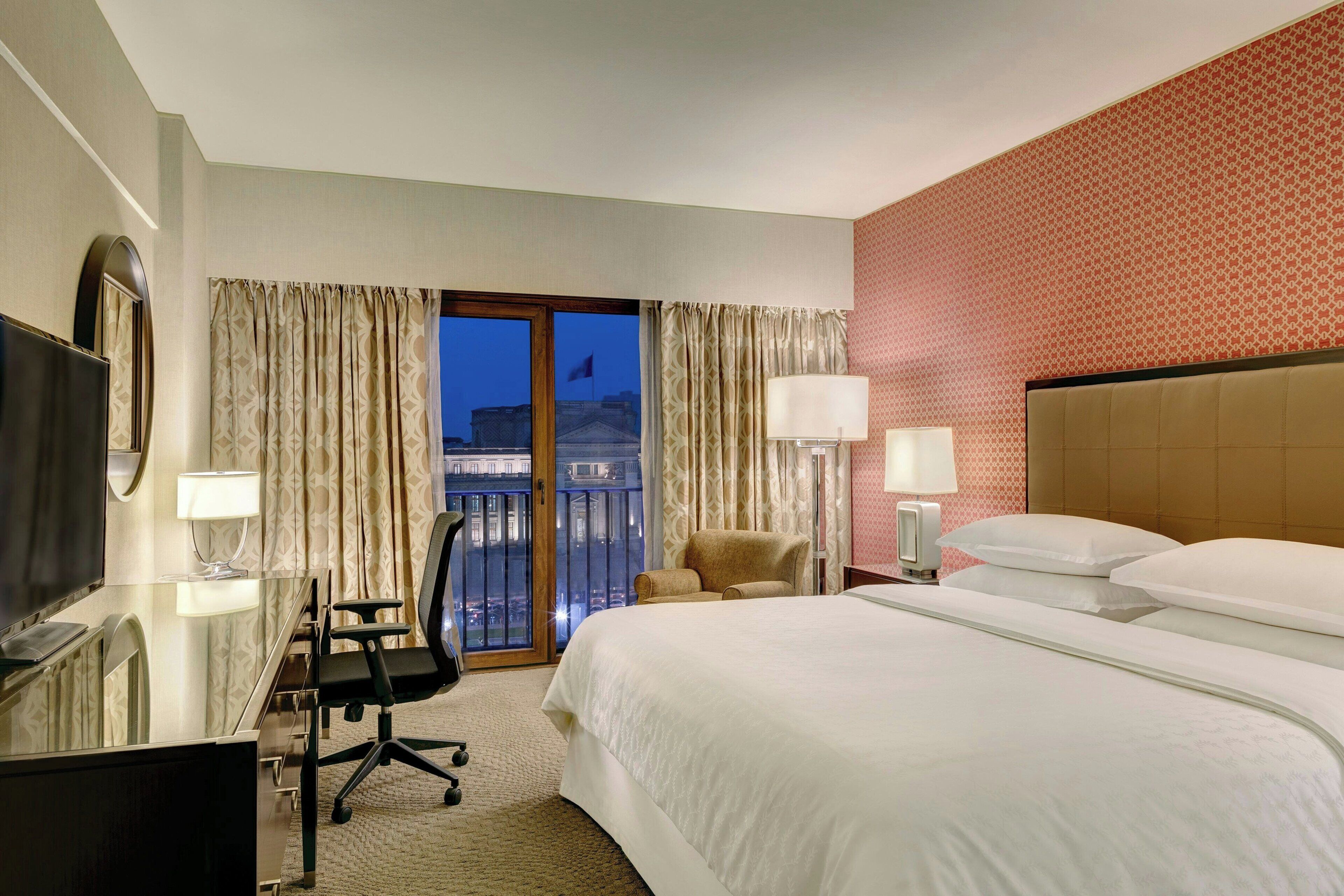 Quarto de hotel com cama, escrivaninha, cadeira e vista para um edifício através de uma janela.
