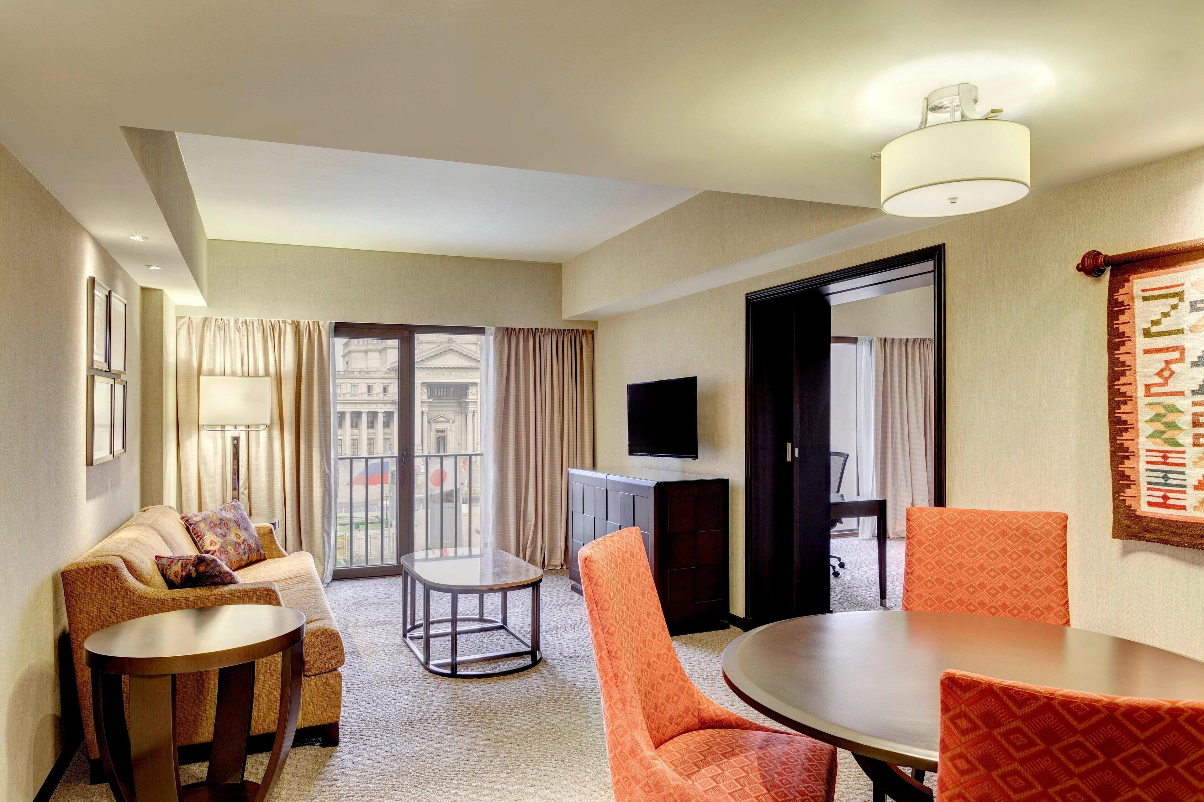Quarto de hotel com sofá, mesa redonda, televisão e varanda com cortinas.