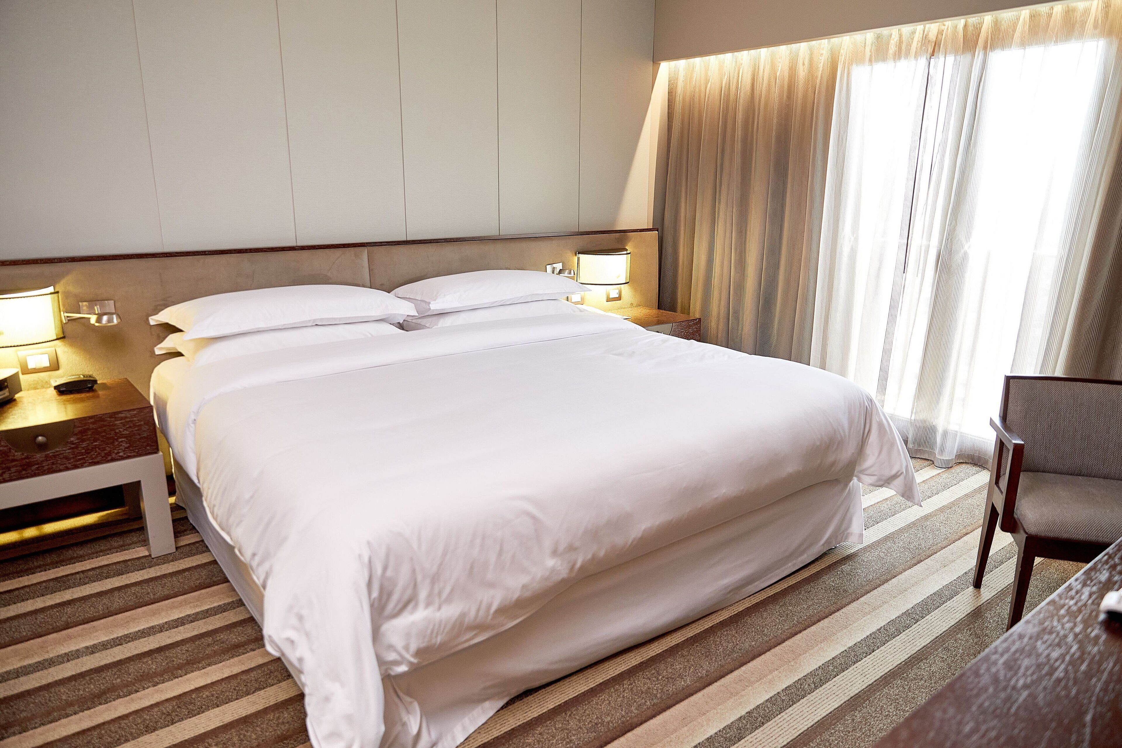 Quarto de hotel com uma cama grande, uma mesa de cabeceira com abajur, uma cadeira e uma janela com cortinas.