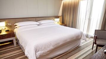 Quarto de hotel com uma cama grande, uma mesa de cabeceira com abajur, uma cadeira e uma janela com cortinas.