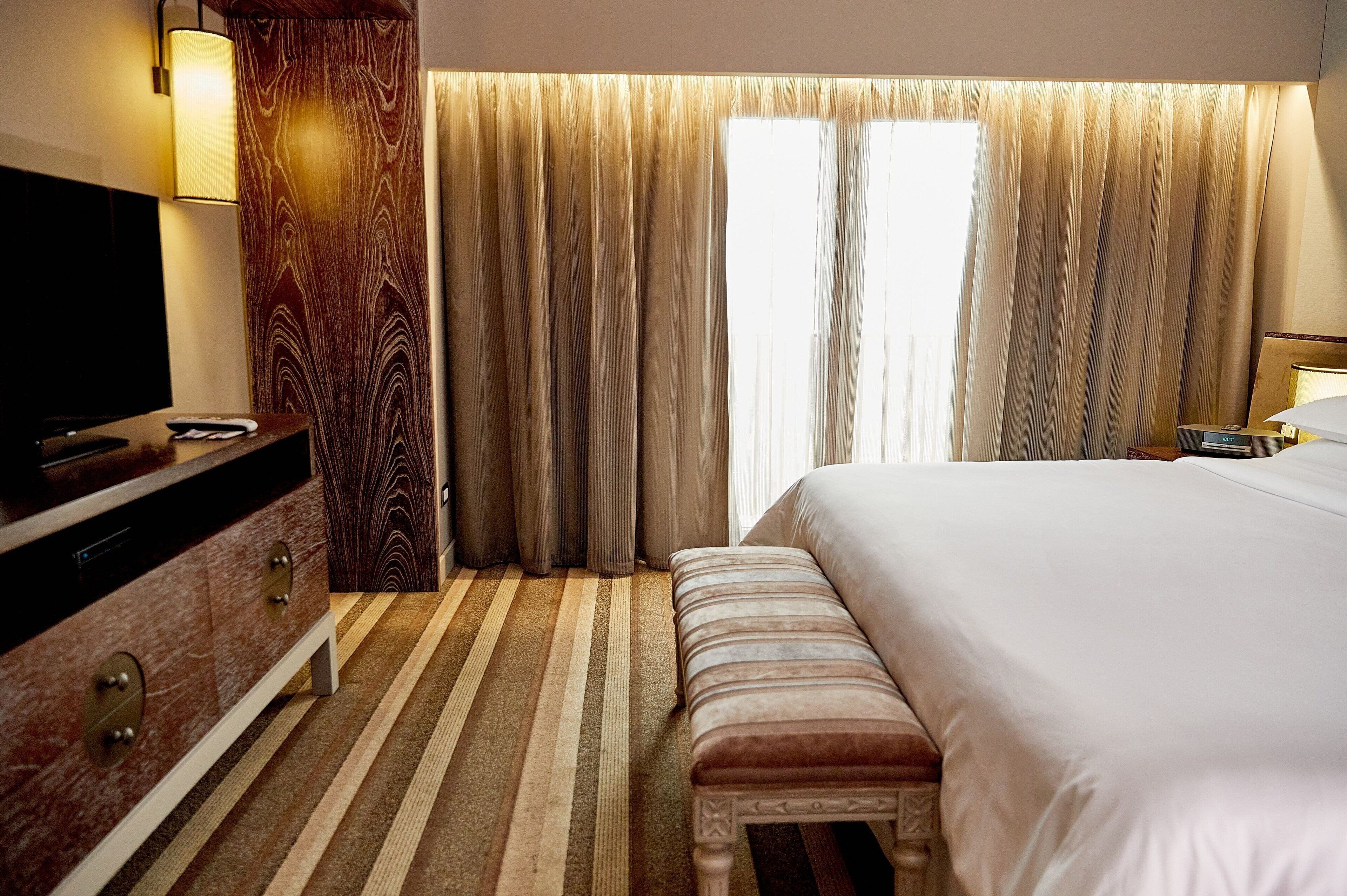 Quarto de hotel com uma cama grande, uma cômoda de madeira com uma televisão e uma janela com cortinas.