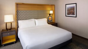 Quarto de hotel com uma cama grande, uma mesa de cabeceira com abajur, um quadro na parede e vista da cidade pela janela.