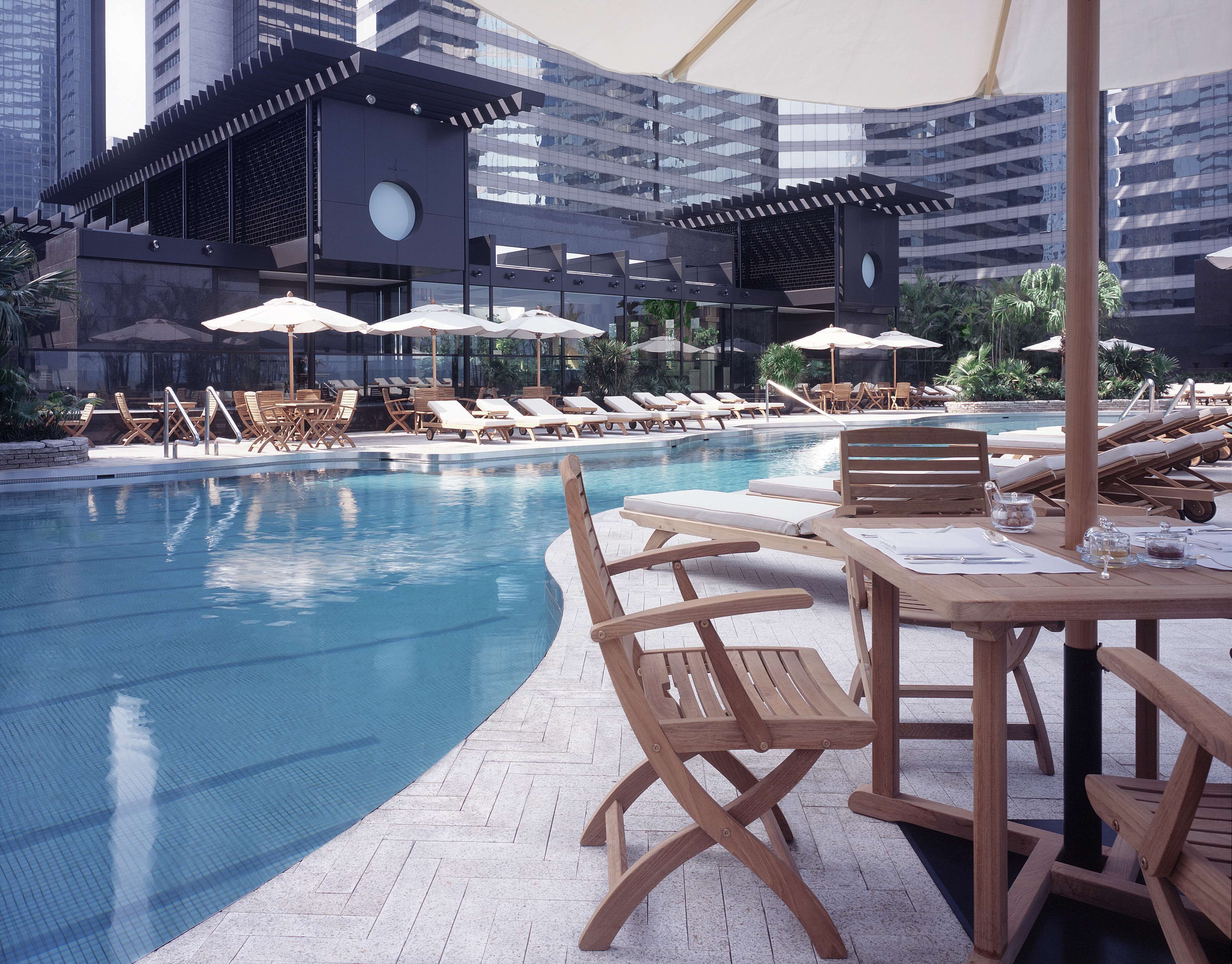 Uma área junto à piscina com espreguiçadeiras e guarda-sóis, rodeada por edifícios modernos.