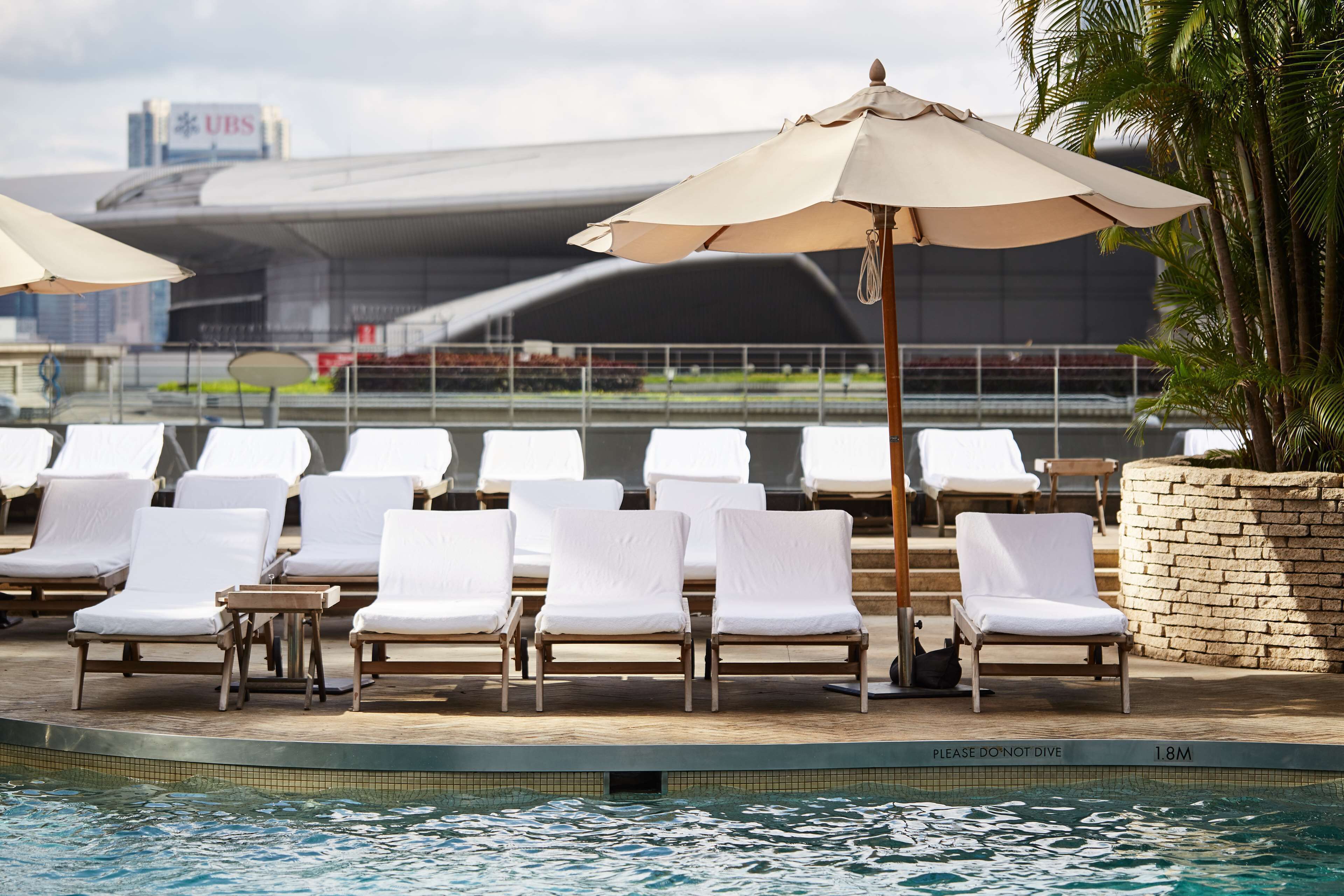 Uma área à beira da piscina com espreguiçadeiras e guarda-sóis, uma piscina límpida e um edifício moderno ao fundo.