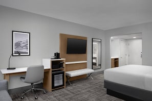 Quarto de hotel com uma cama, uma secretária com cadeira, televisão, pequeno armário e um quadro na parede.