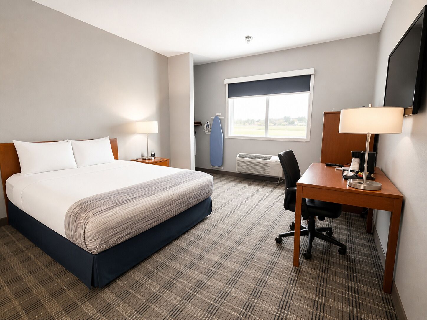 Quarto de hotel com uma cama, uma escrivaninha, uma cadeira, uma televisão e uma janela.