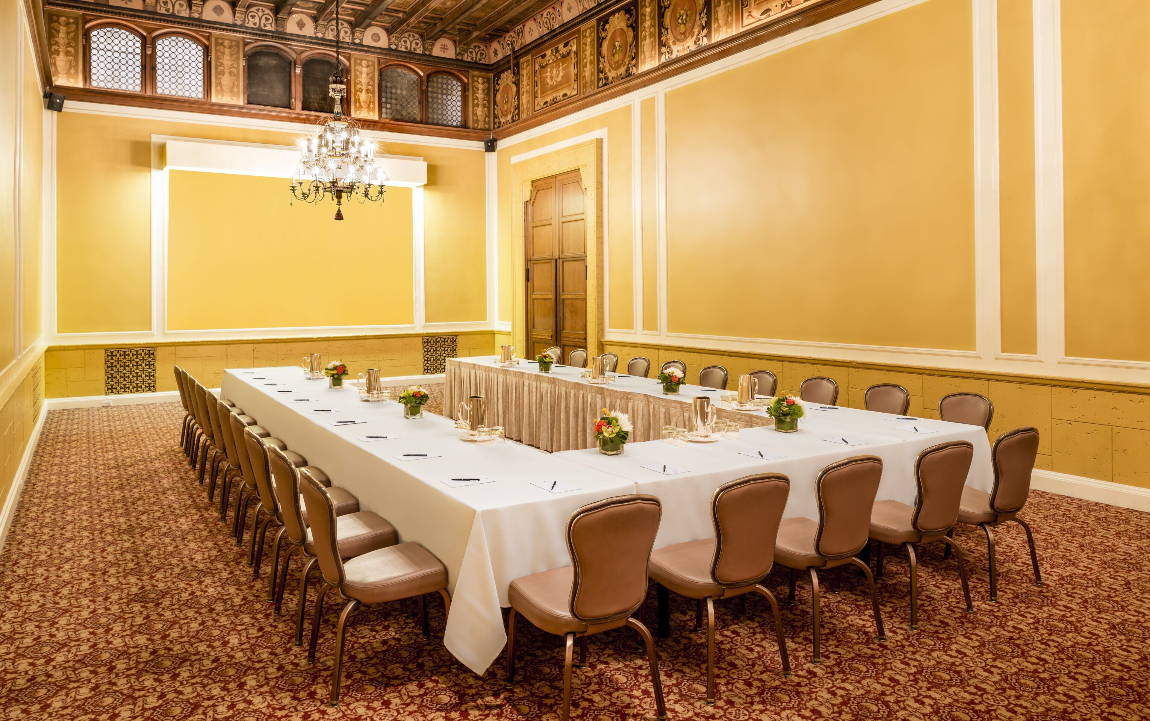 Uma sala de conferências com uma mesa comprida, cadeiras, um candelabro e detalhes ornamentais no teto.