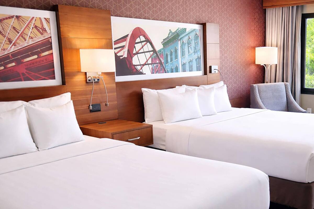 Quarto de hotel com duas camas, uma mesa de cabeceira, um abajur e duas pinturas penduradas na parede.