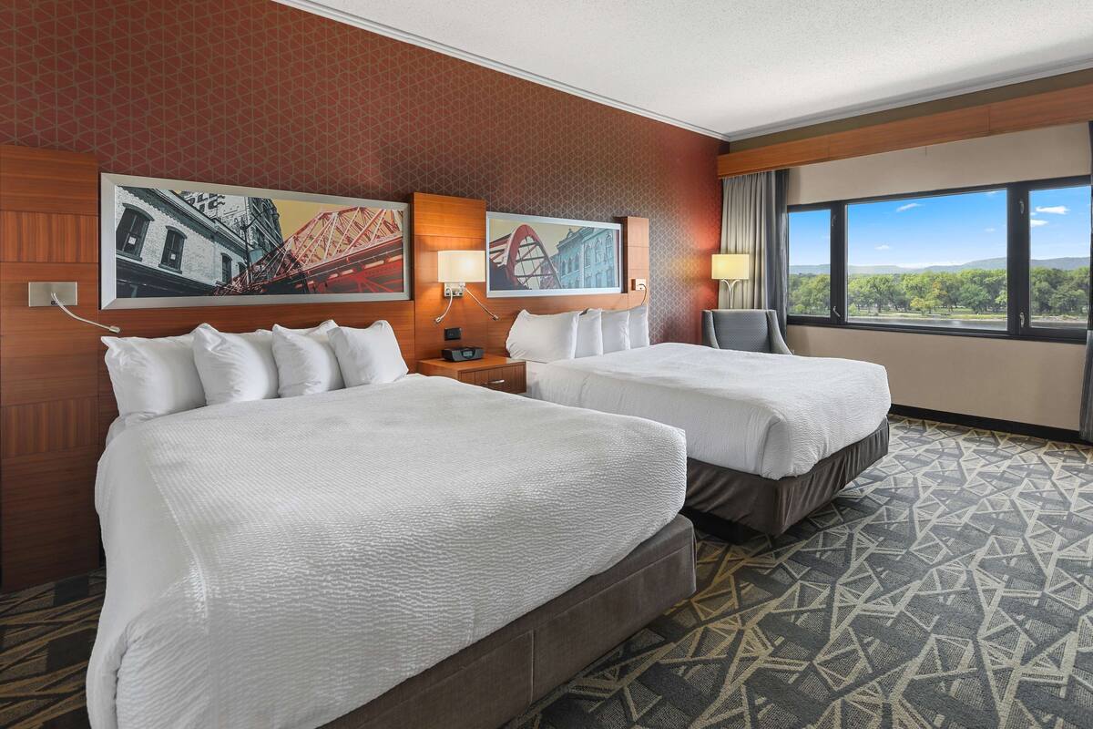 Quarto de hotel com duas camas, uma janela grande com vista e duas pinturas na parede.