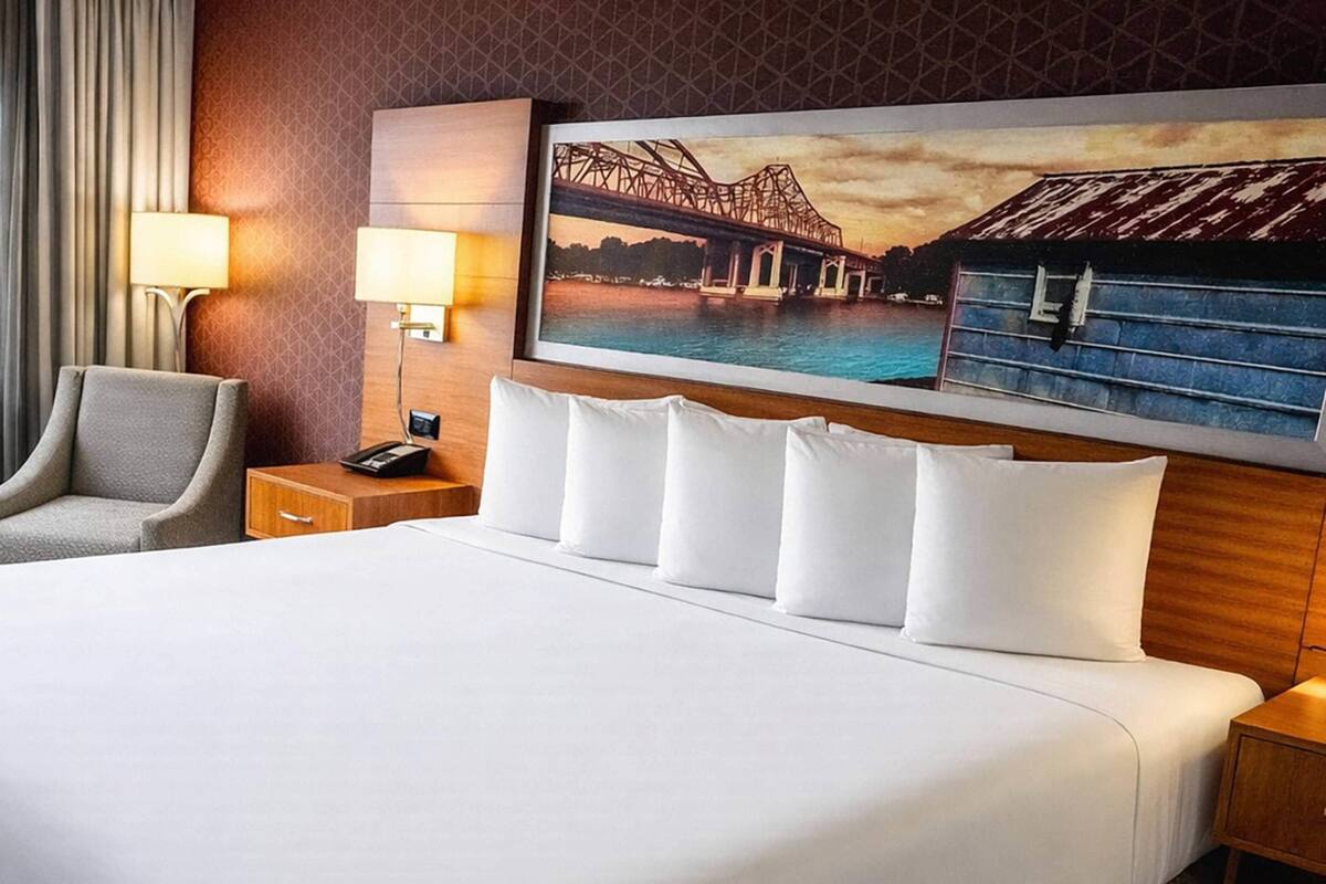 Quarto de hotel com uma cama grande, uma cadeira, uma mesinha de cabeceira, um abajur e um quadro de uma ponte.