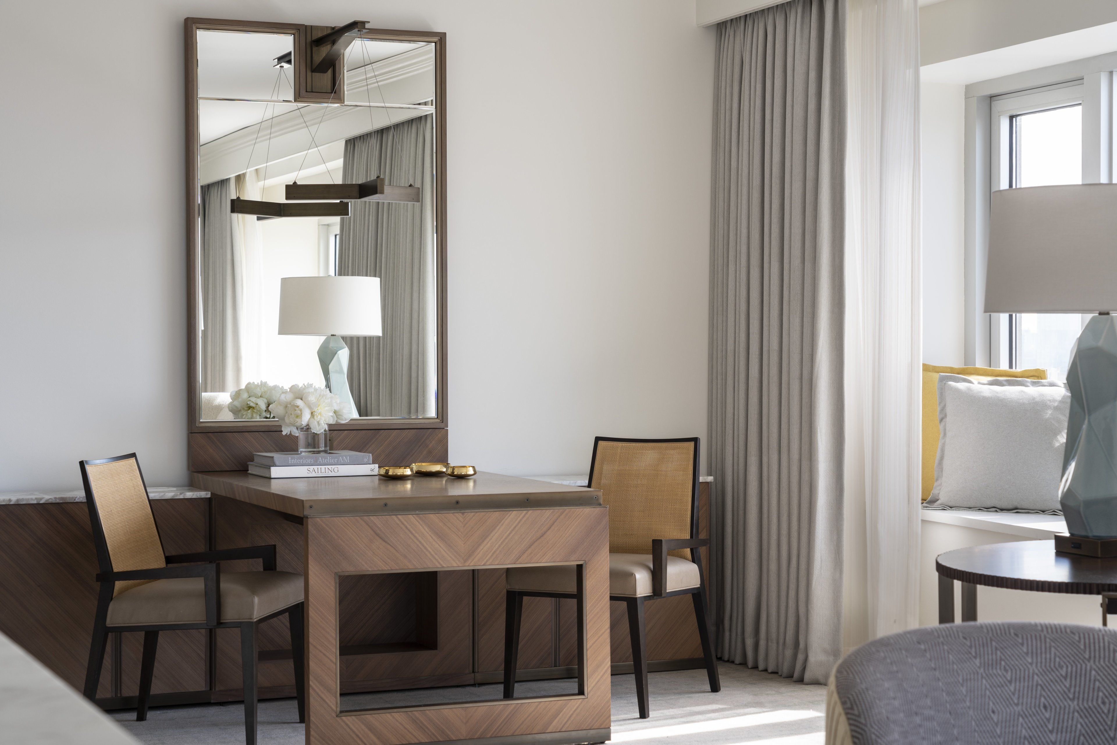 Quarto de hotel com escrivaninha de madeira, duas cadeiras, um espelho grande, abajur e janela com cortinas.